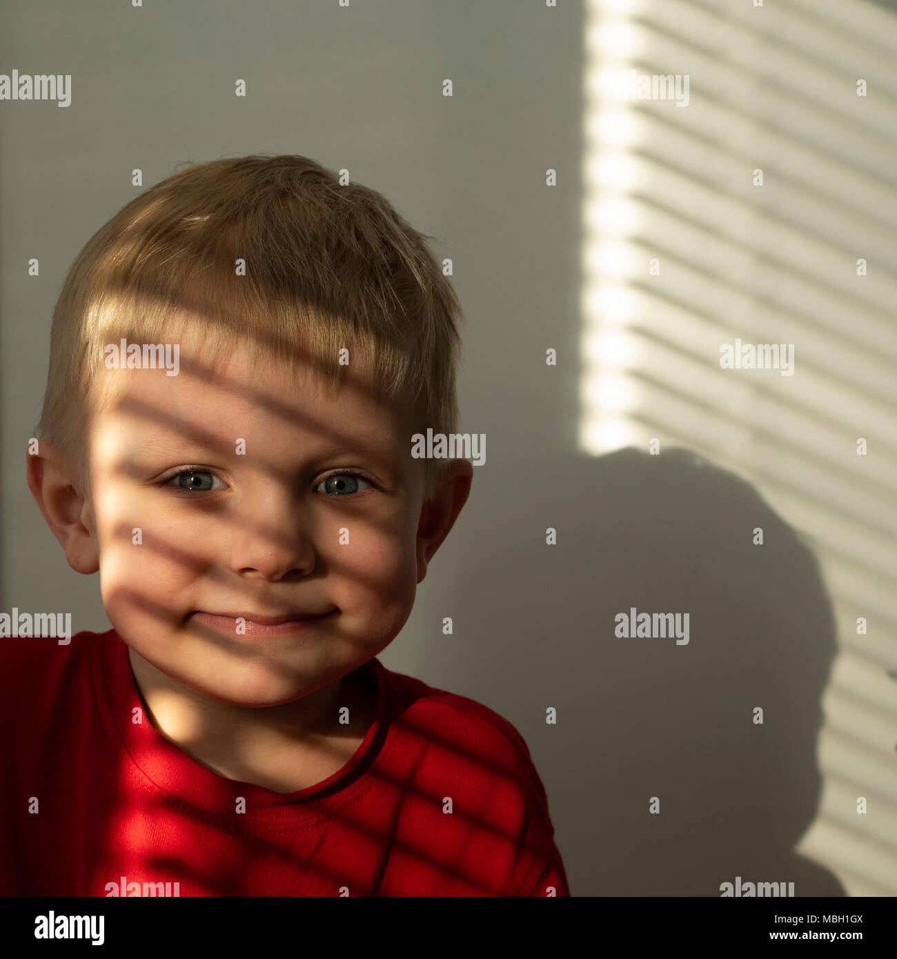 Smiling caucasian boy in a red t-shirt with a shadow from the blinds ...