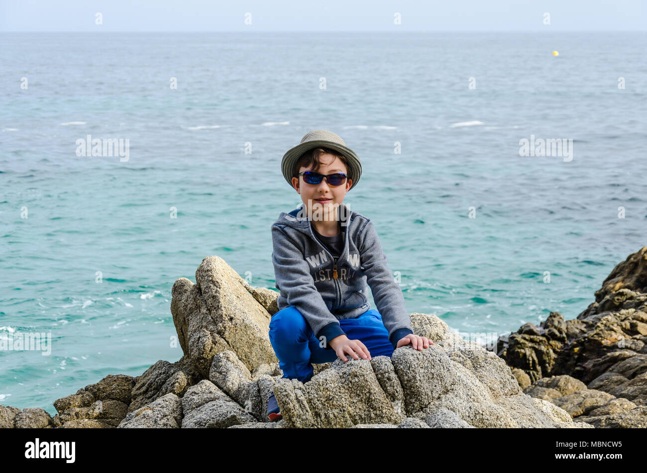 A young boy crouches down on rocks near the sea and poses for a ...