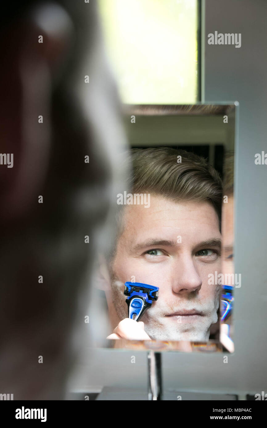 Reflection of handsome man with green eyes shaving with blue razor in ...
