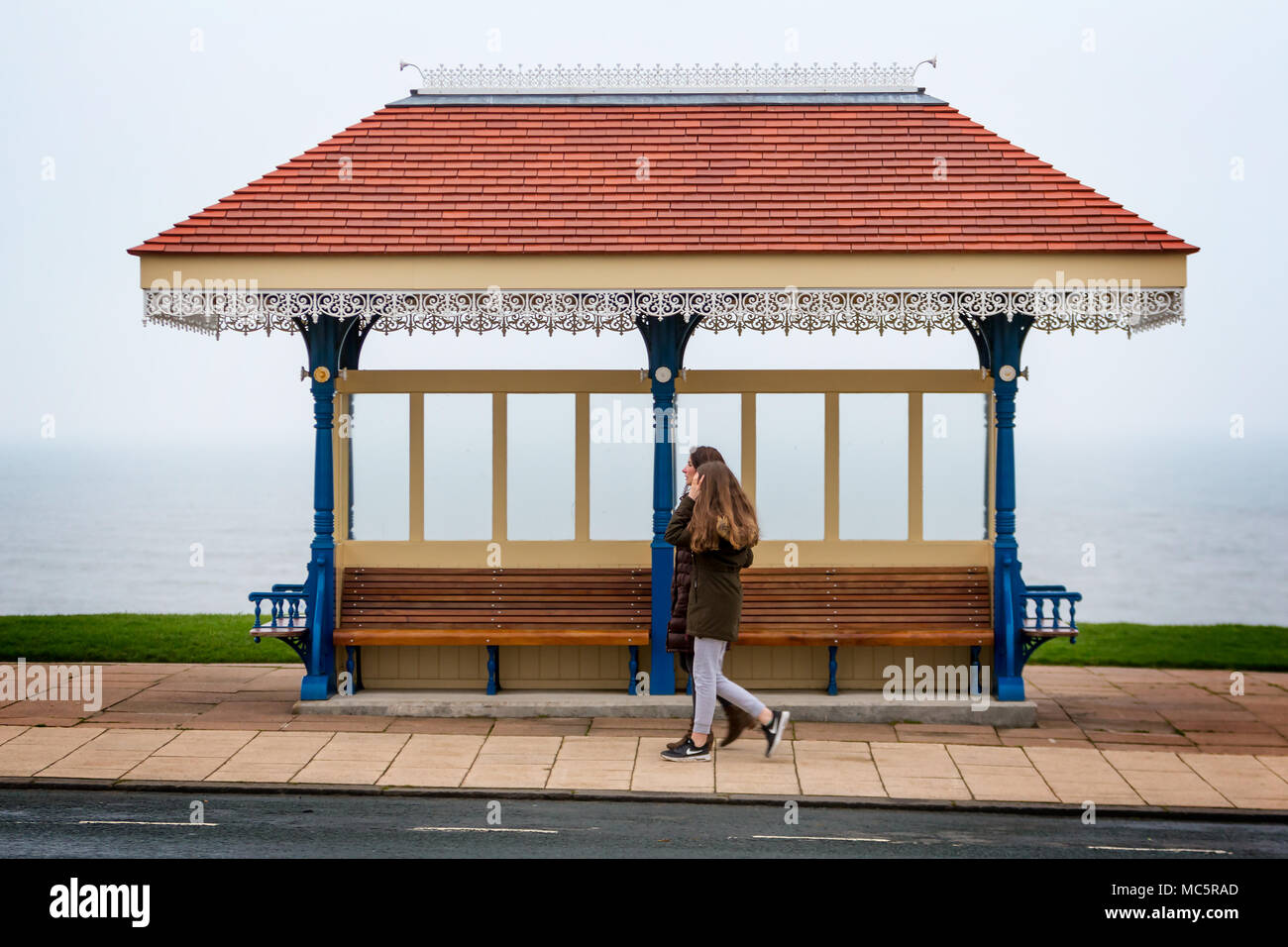 Two people walk along the top of Whitby's West Cliff area past a seated ...