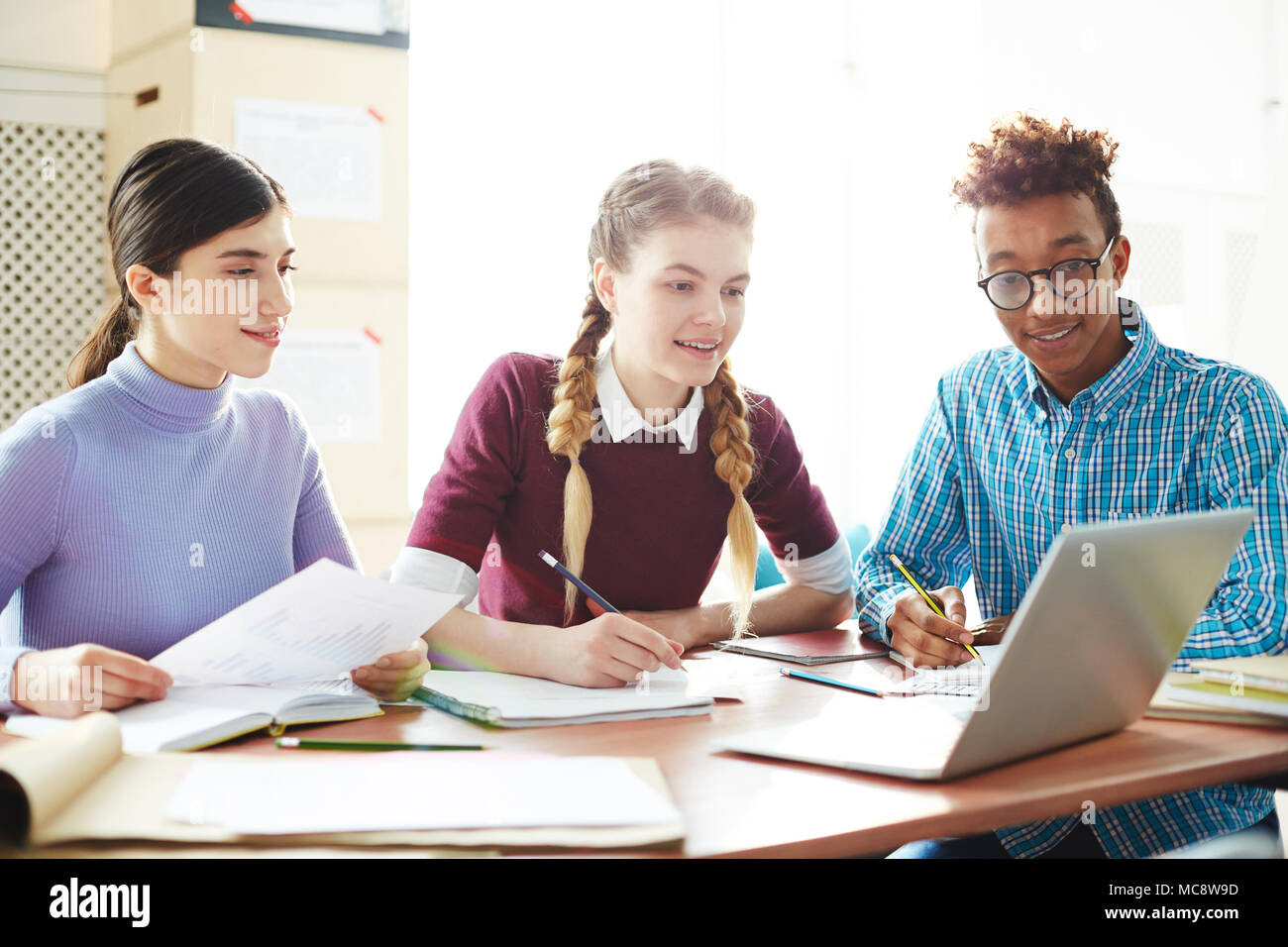 Group of clever students sitting by desk in front of laptop and reading ...