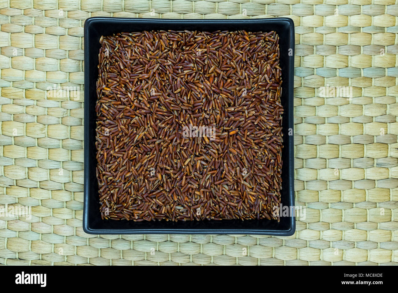 Red rice grains in black plate on natural bamboo mat background surface ...