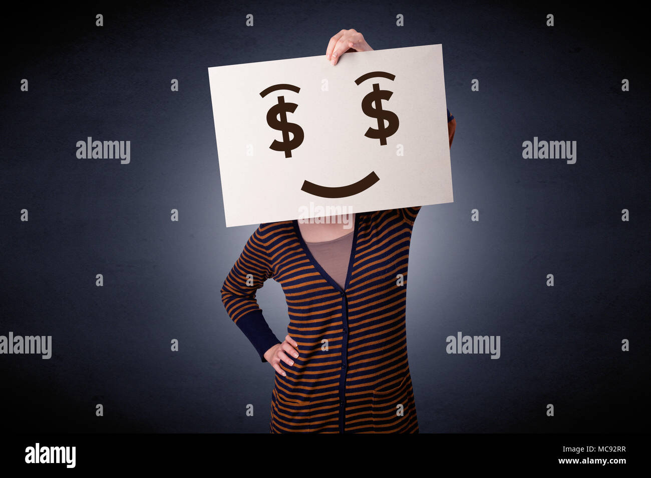 Young casual woman hiding behind a greedy face drawing on cardboard ...