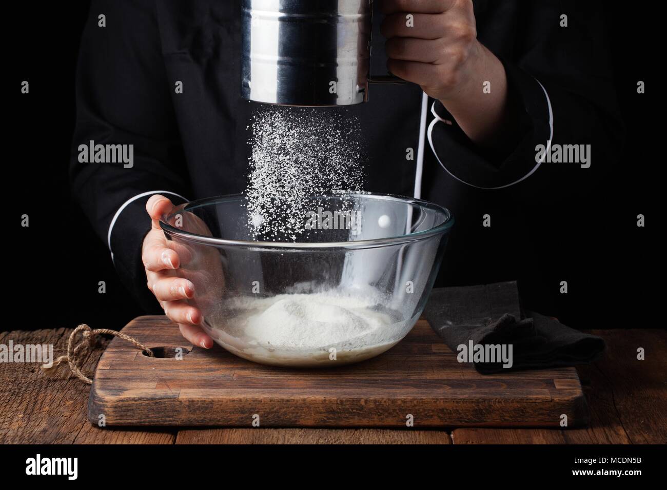 Chef hands pouring flour powder on raw dough using sieve on a black ...