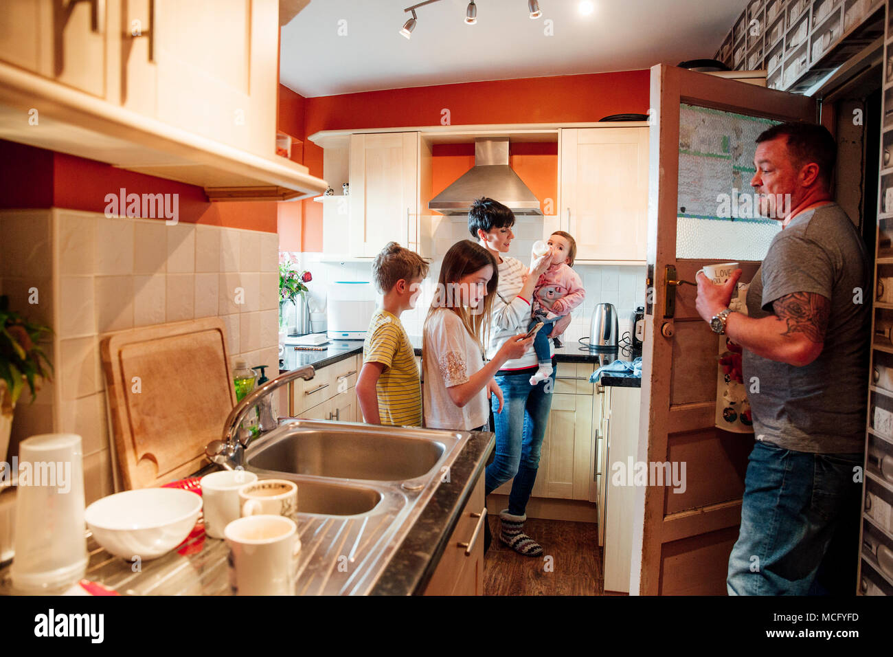 A whole family are busy in the kitchen of their home. The mother is ...