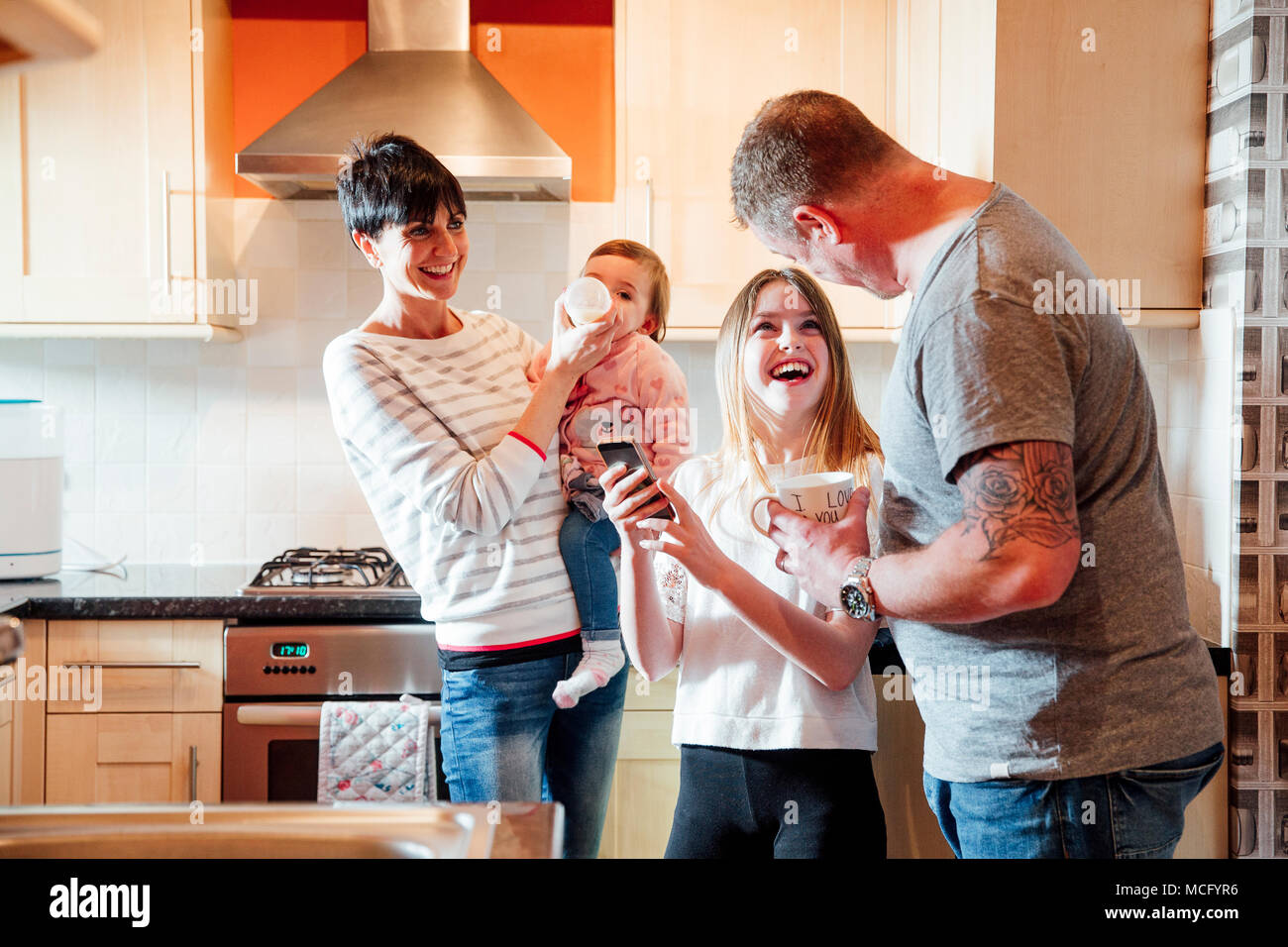 A family are socialising in the kitchen. the mother is feeding the baby ...