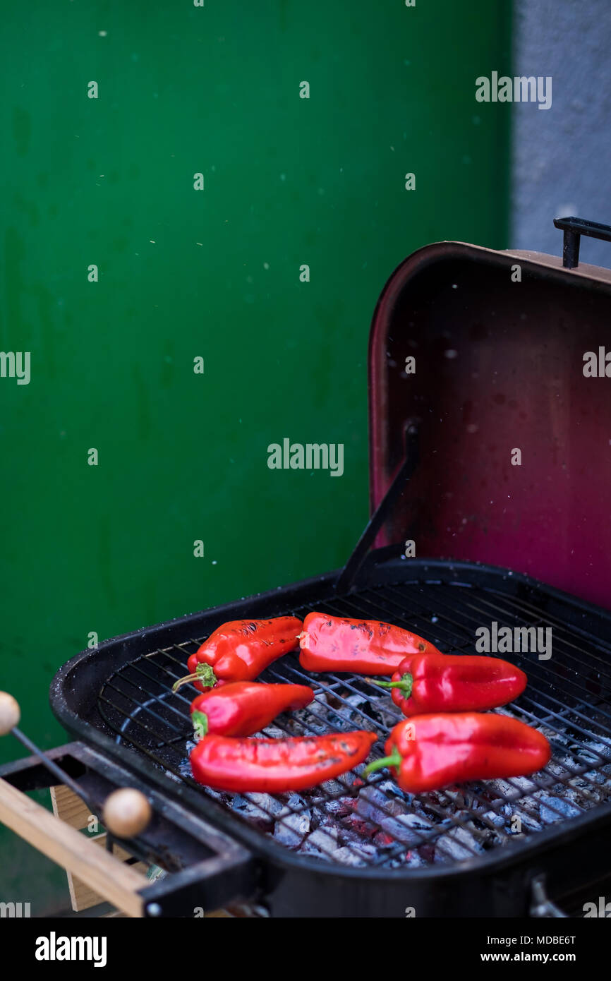 Red peppers roasting on a charcoal barbecue outside near a green wall ...