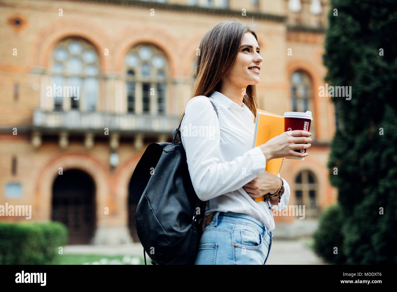 Smiling young student girl holding coffee cup, outdoors with notebooks ...