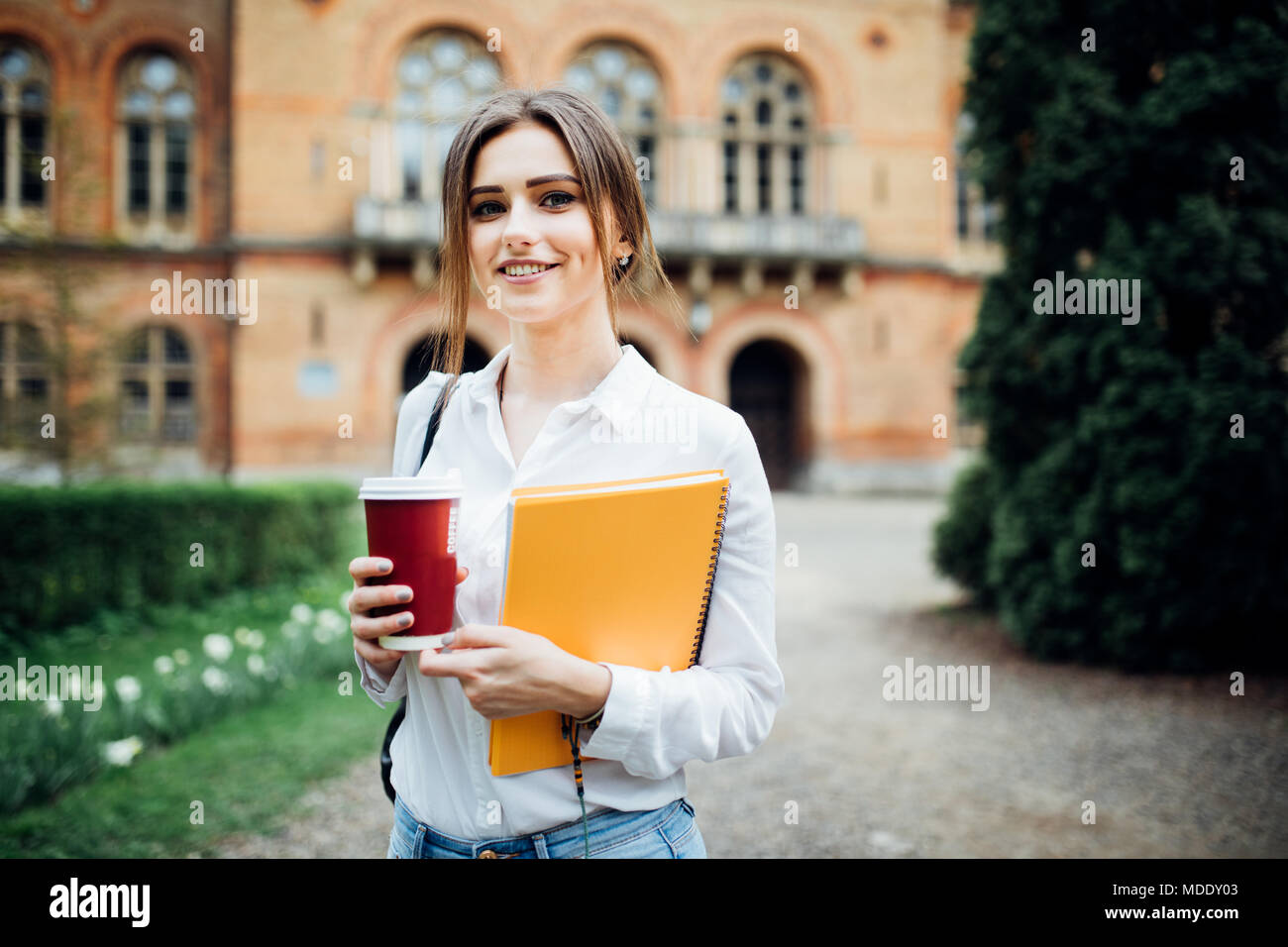 Smiling young student girl holding coffee cup, outdoors with notebooks ...