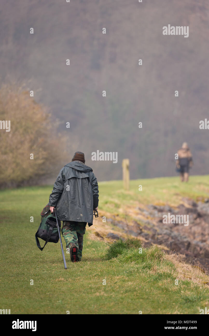 Rear view of UK fisherman in waterproofs carrying tackle: fishing rod ...