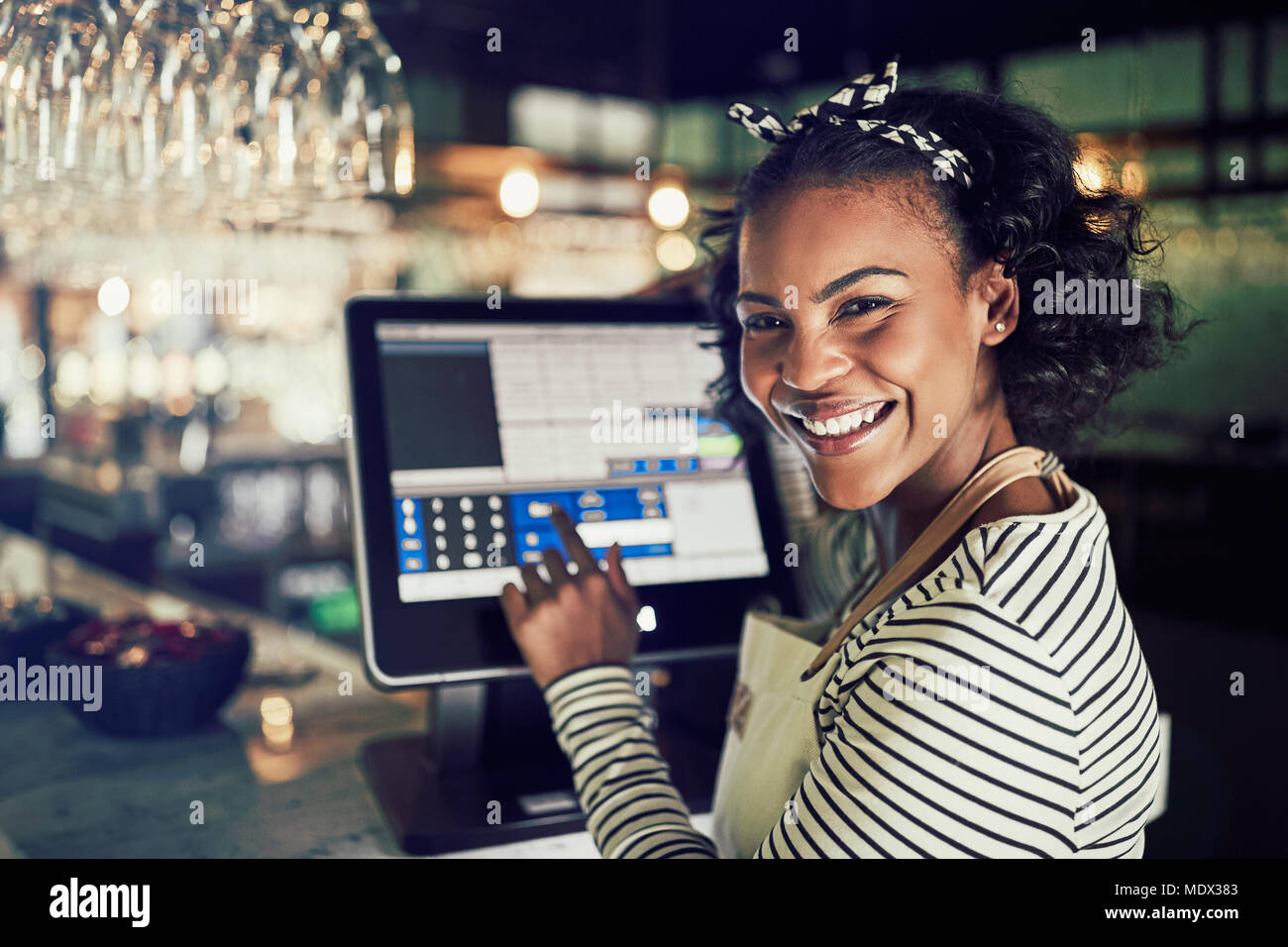 Smiling young African waitress wearing an apron using a touchscreen ...