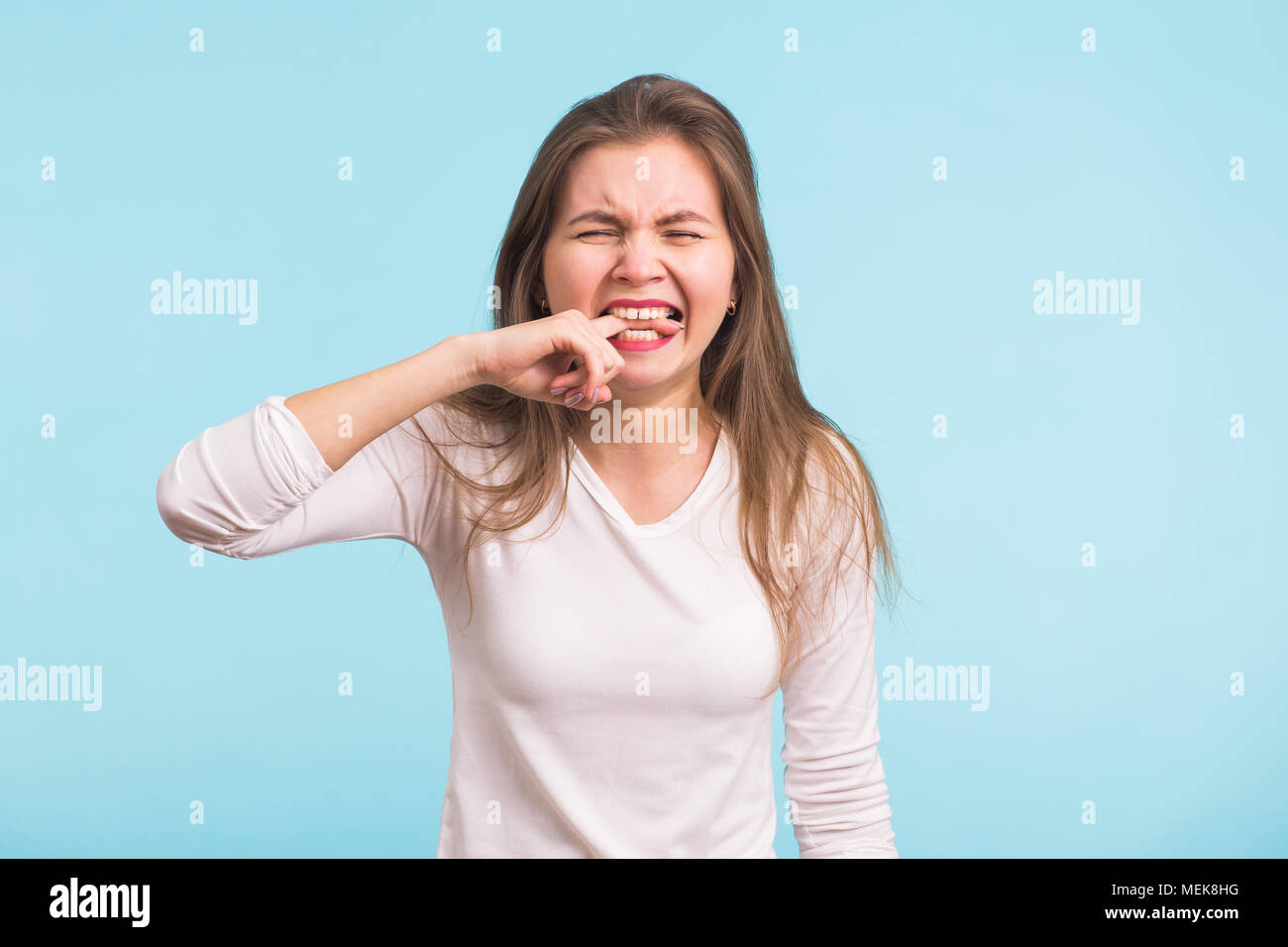 Woman biting finger with her teeth, expresses pain and anger on blue ...