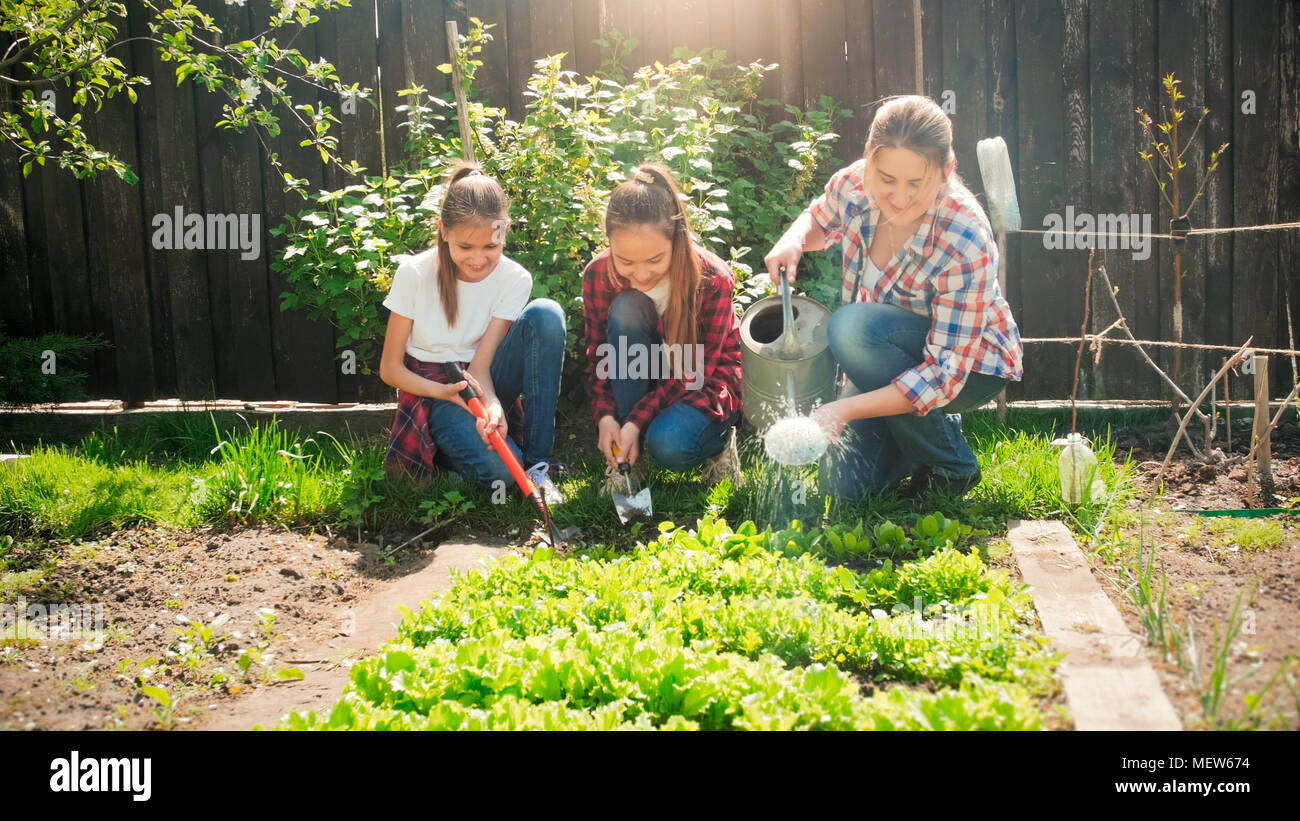 Two teenage girls weeding garden bed while young mother watering ...