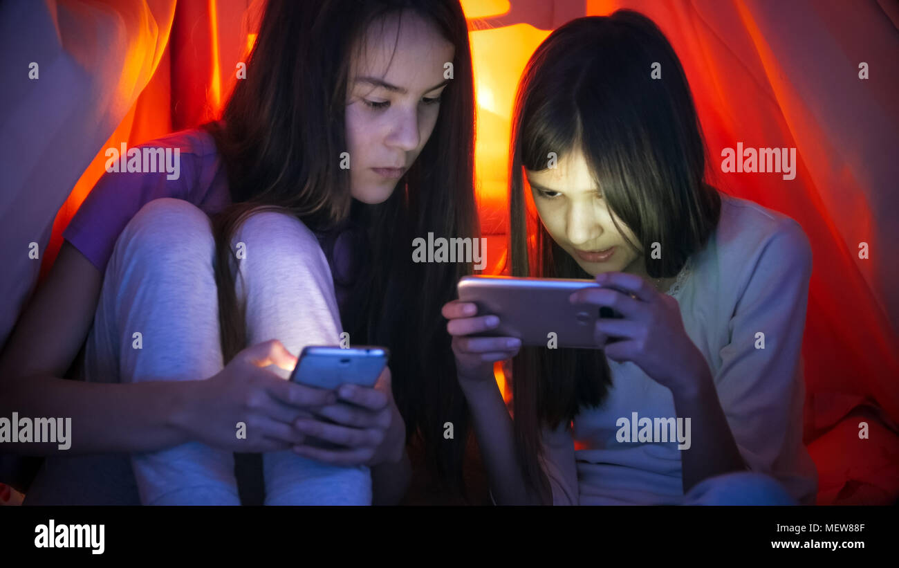 Portrait of two teenage girls with smart phones under blanket at night ...