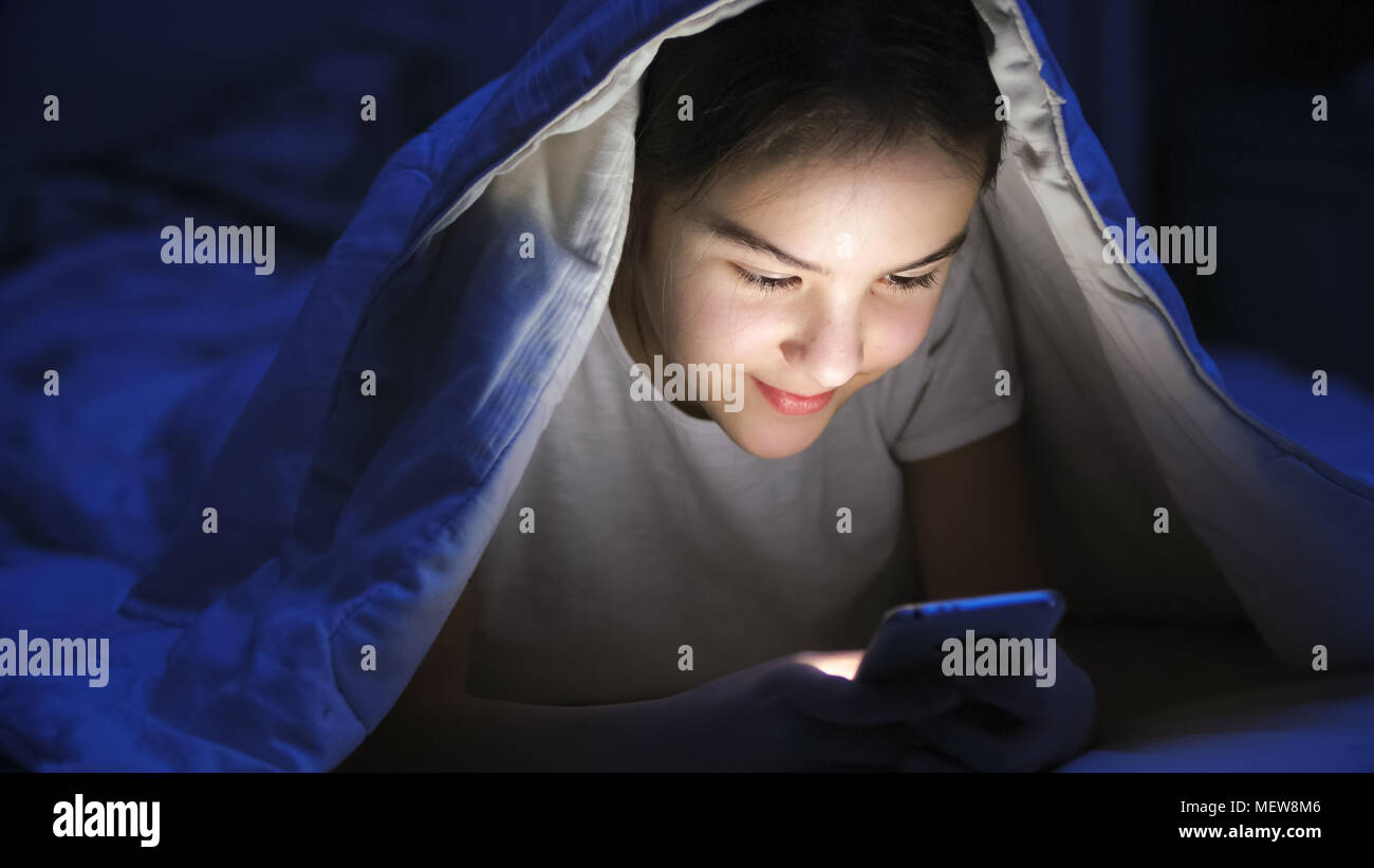 Portrait of teenage girl typing message on smartphone under blanket at ...