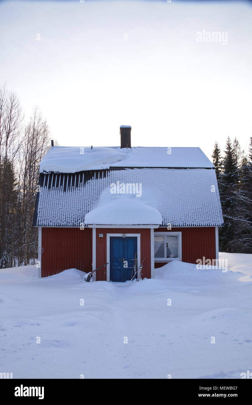 A small red wooden cottage is covered in deep snow on a cold winter day ...