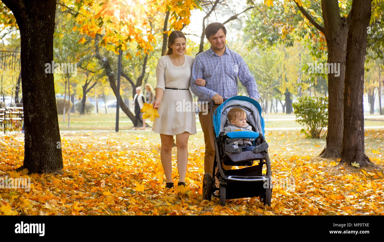 Happy smiling family walking with toddler son sitting in pram at autumn ...