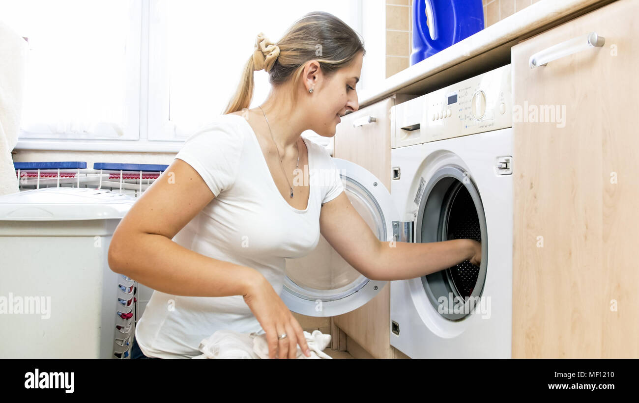Portrait of young smiling woman loading clothes in washing machine ...