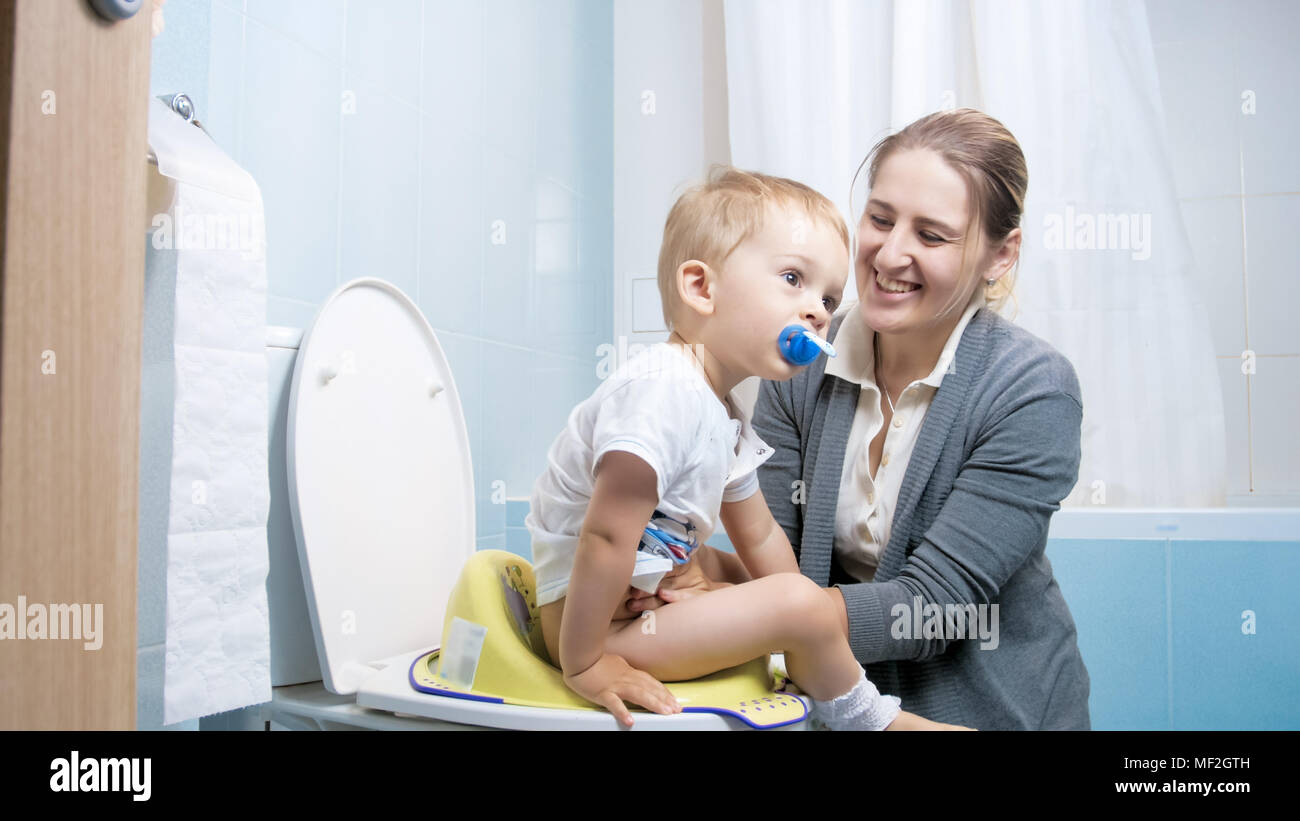 Portrait of smiling young mother teaching her toddler son using toilet ...