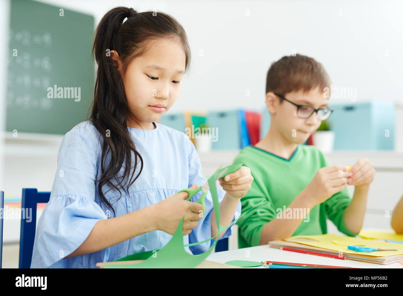 Little Asian girl and her Caucasian classmate cutting colored paper ...