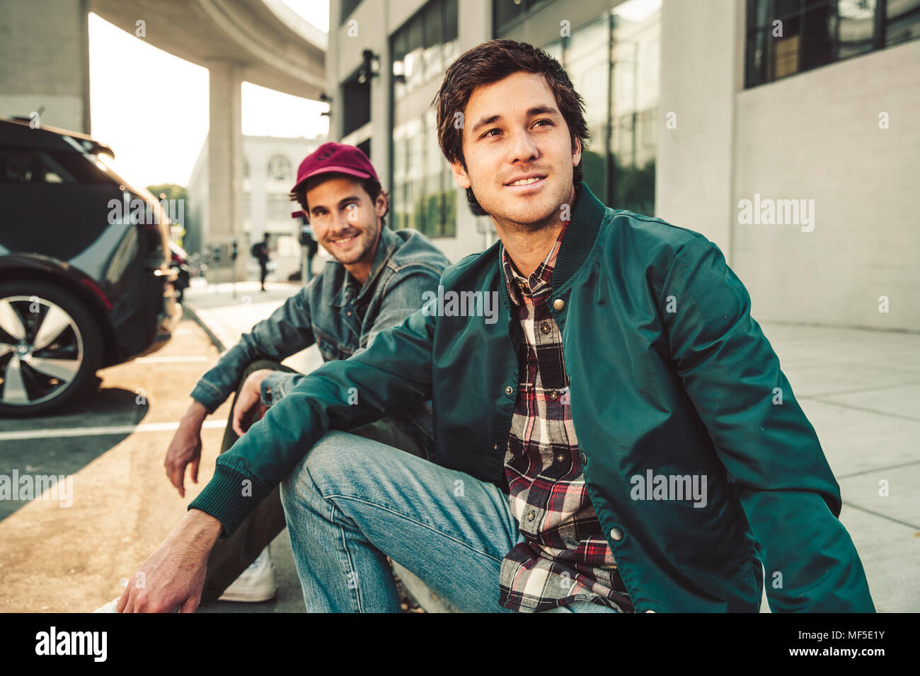 Two smiling young men sitting on sidewalk Stock Photo - Alamy