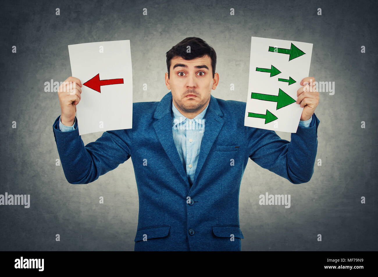 Confused young businessman holding two sheets with drawn arrows poinded ...