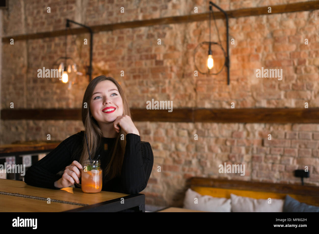 Satisfied girl sitting near wooden table with cup of lemonade in cafe ...
