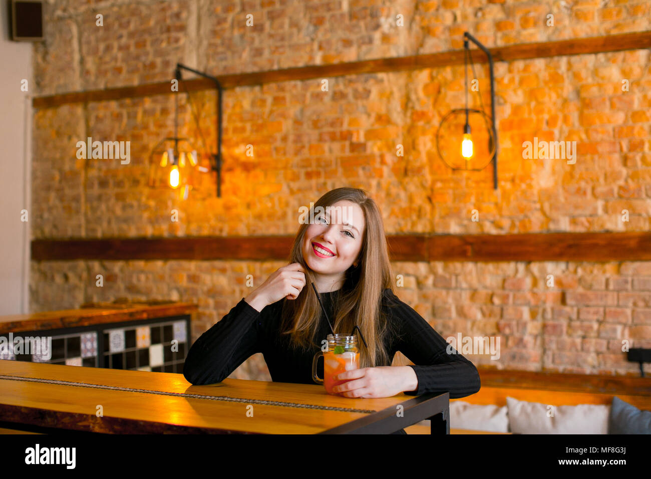 Satisfied girl sitting near wooden table with cup of lemonade in cafe ...