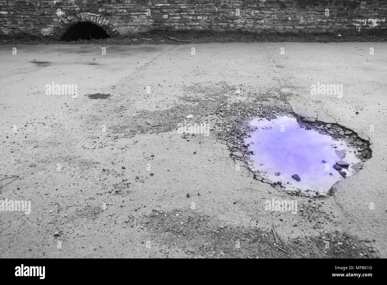 pothole in UK road with bright blue water and archway in background ...