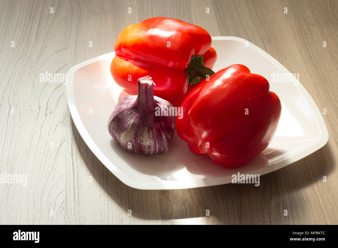 Two red bell peppers and a head of blue garlic lie on a white plate ...