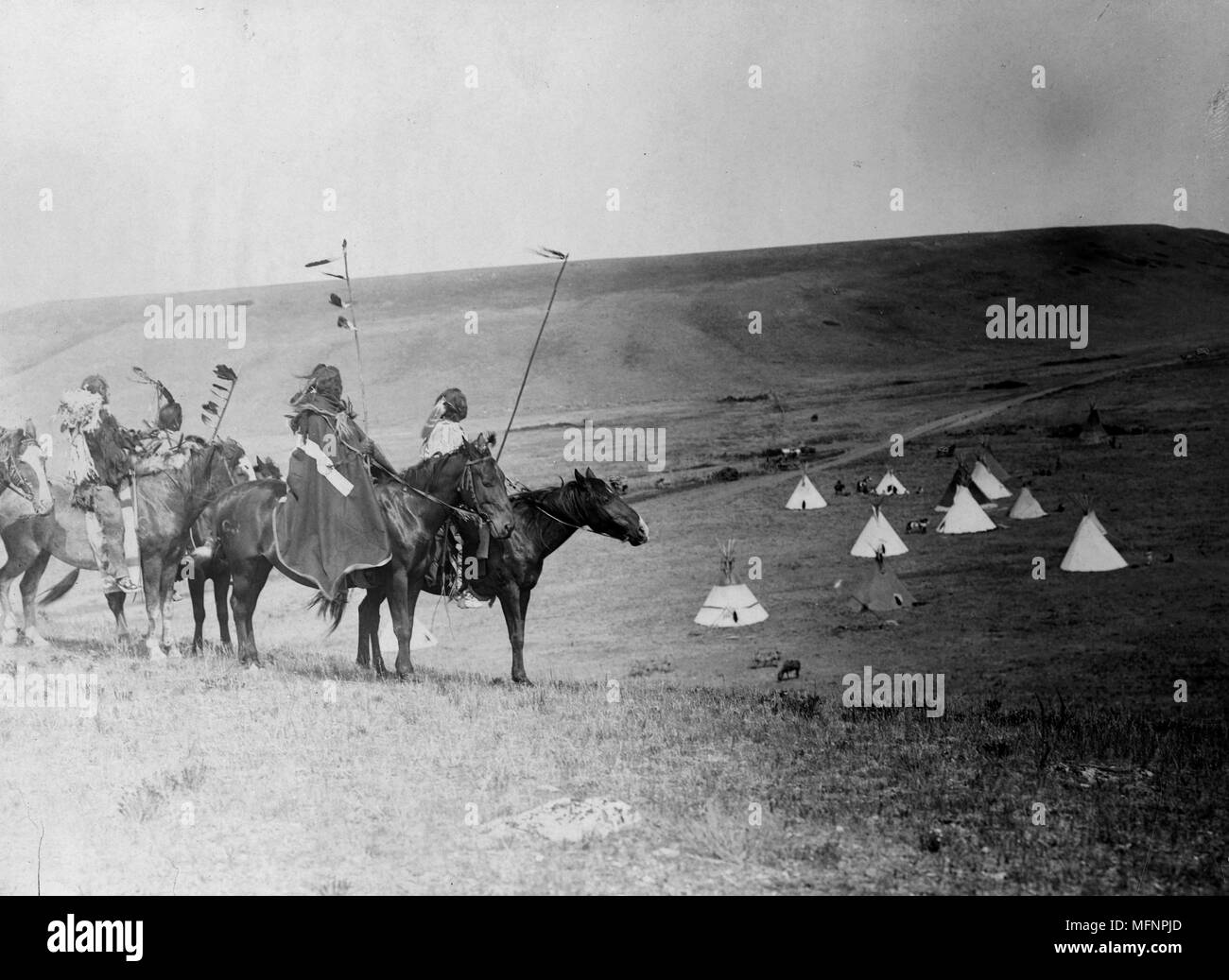 Four Atsina Indians on horseback overlooking tepees in valley beyond ...