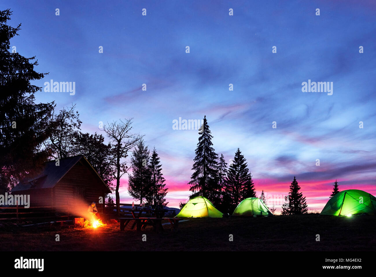 Night camping. Tourist have a rest at a campfire near illuminated tent ...