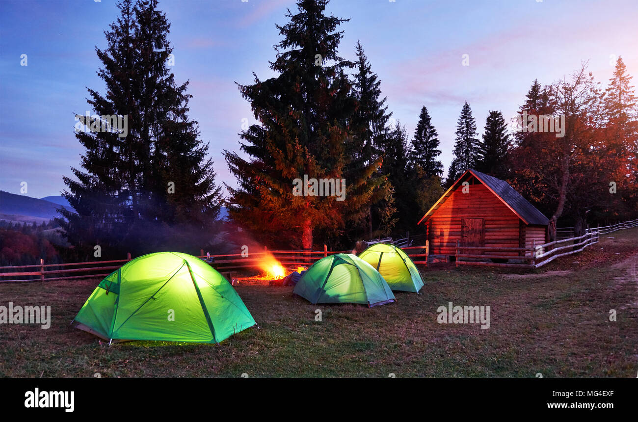 Night camping. Tourist have a rest at a campfire near illuminated tent ...