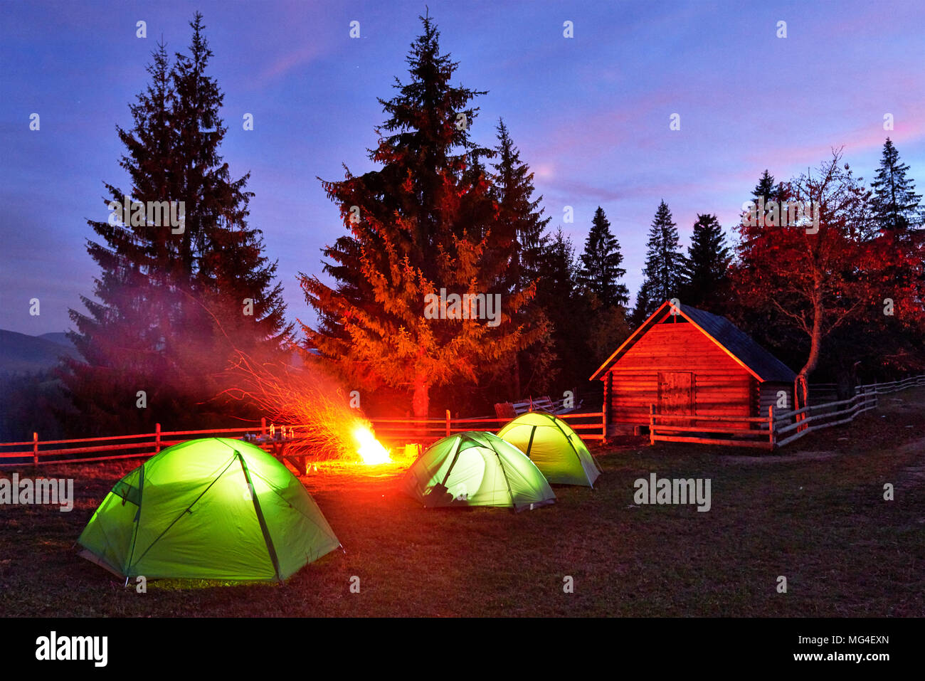 Night camping. Tourist have a rest at a campfire near illuminated tent ...