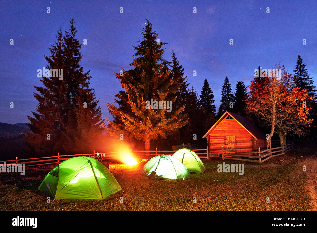 Night camping. Tourist have a rest at a campfire near illuminated tent ...