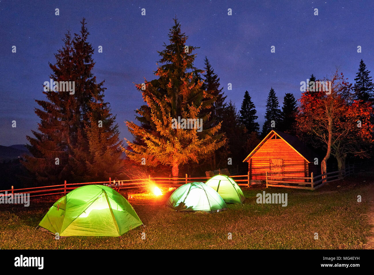 Night camping. Tourist have a rest at a campfire near illuminated tent ...