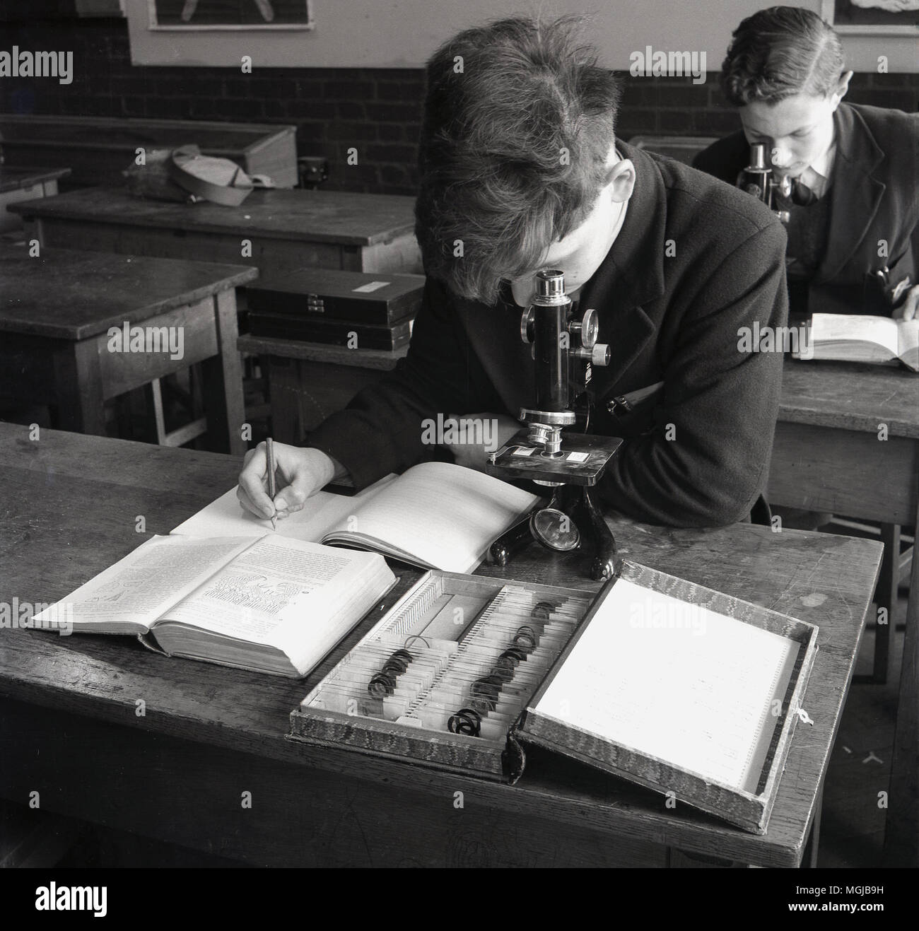 1950s, historical, schoolboy at a desk using a microscope and taking ...