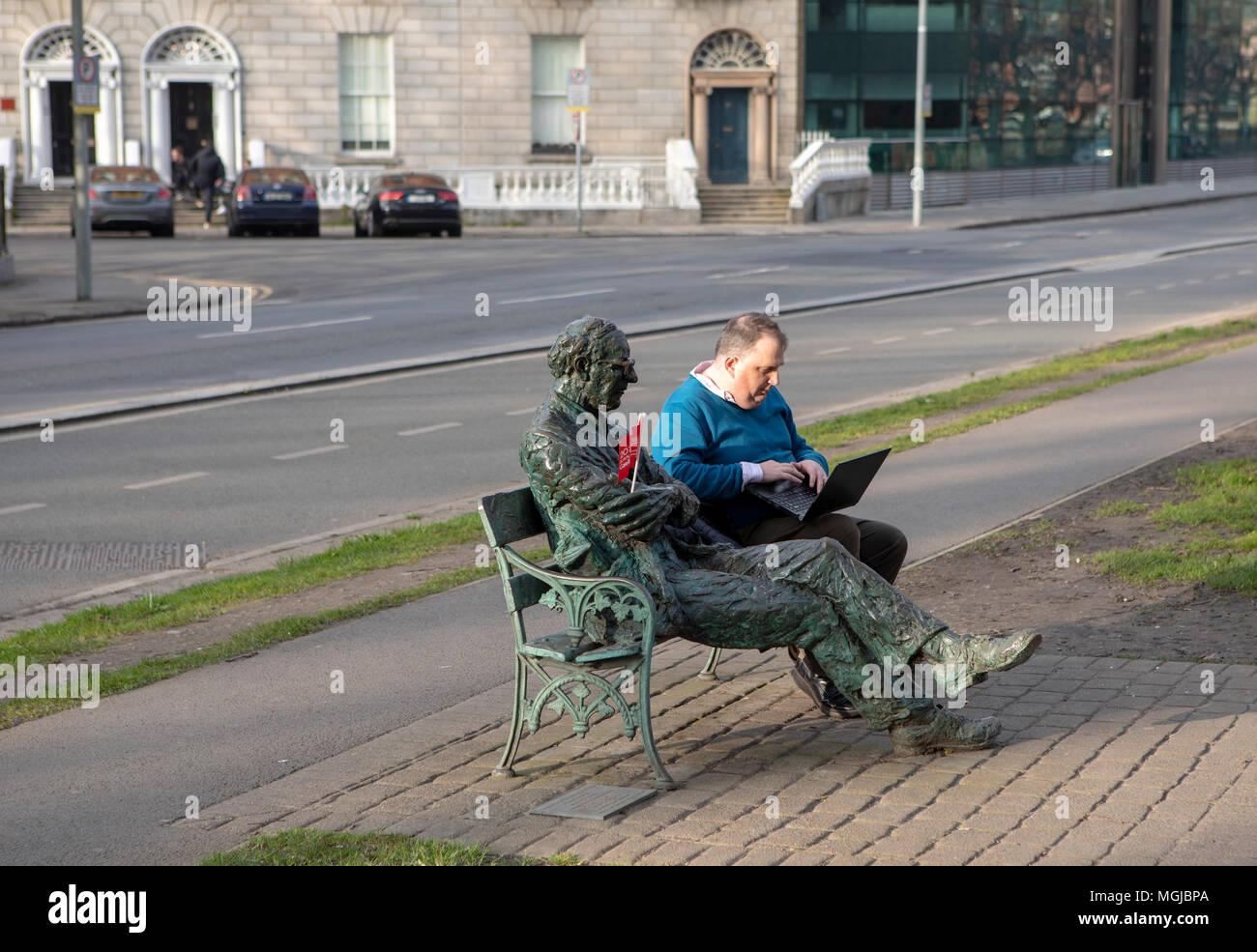 Man works outdoors on bench with laptop beside statue of Irish poet ...
