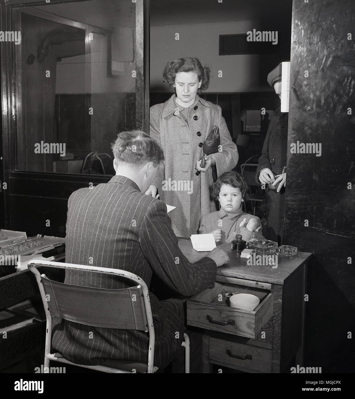 1950s, mother with her young daughter hands over paperwork to a male ...