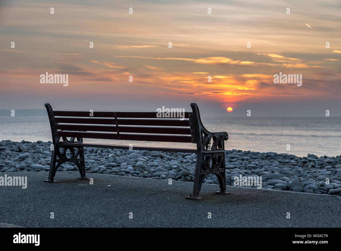 An empty bench, at the sea shore, with a fiery, colorful sun setting ...