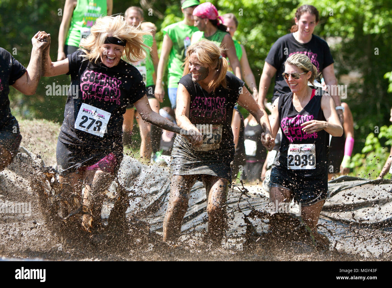 Women laugh and hold hands as they run and splash through a mud pit at ...
