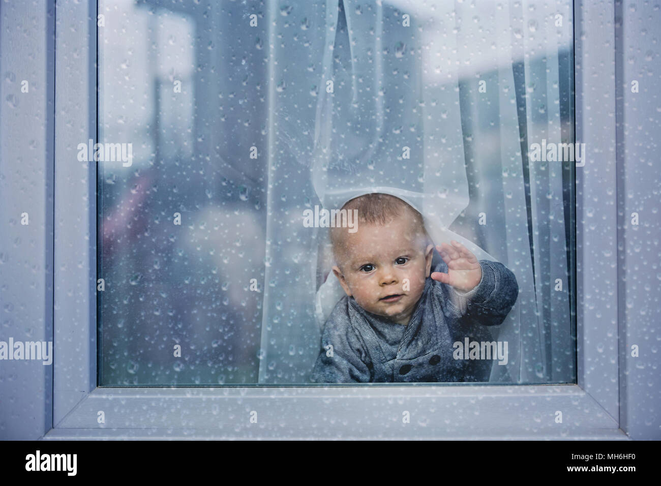 Cute toddler boy, staying at home behind the glass doors or window ...