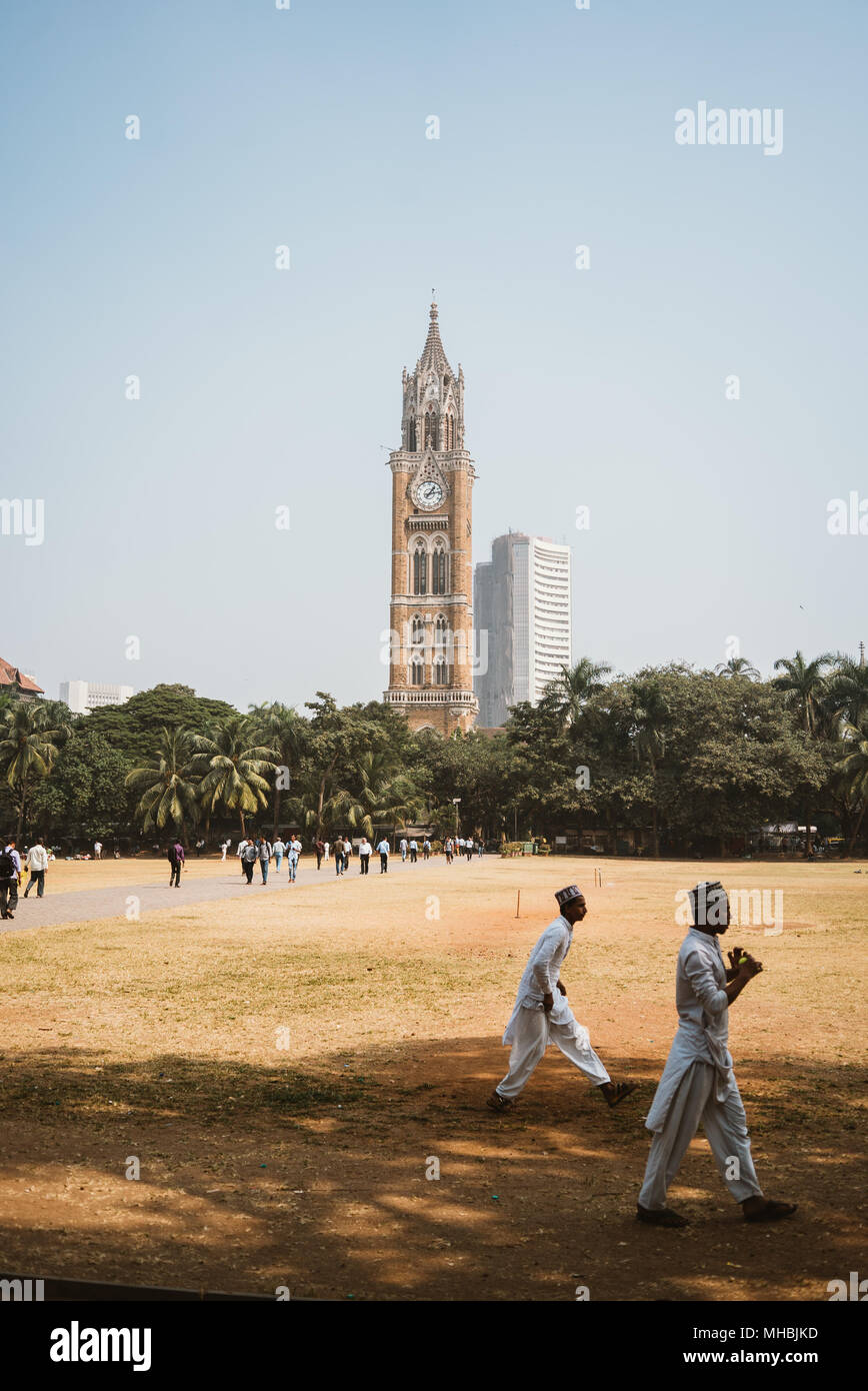 Indian boys playing cricket in a dry field with a gothic clock tower in ...