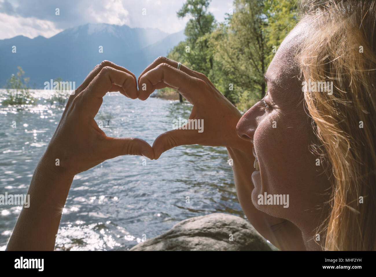 Young woman by the lake making heart shape with hands on mountain ...