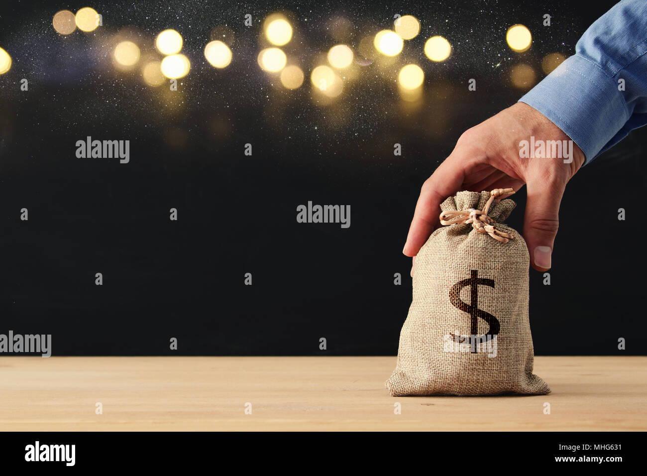 Male hand holding a sack of money over wooden desk and glitter black ...