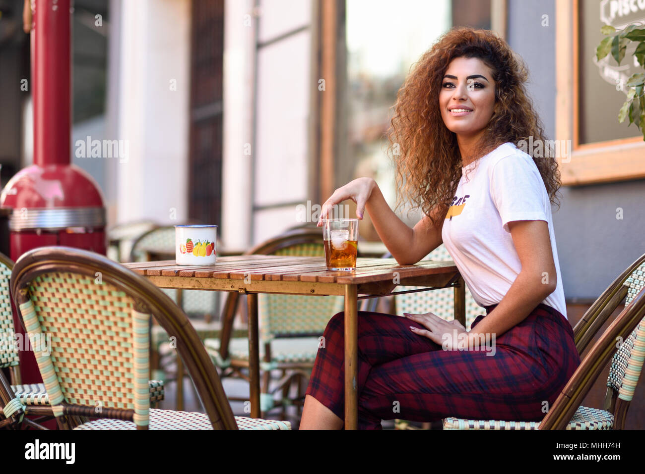 Young arabic woman smiling and sitting in an urban bar in the street ...