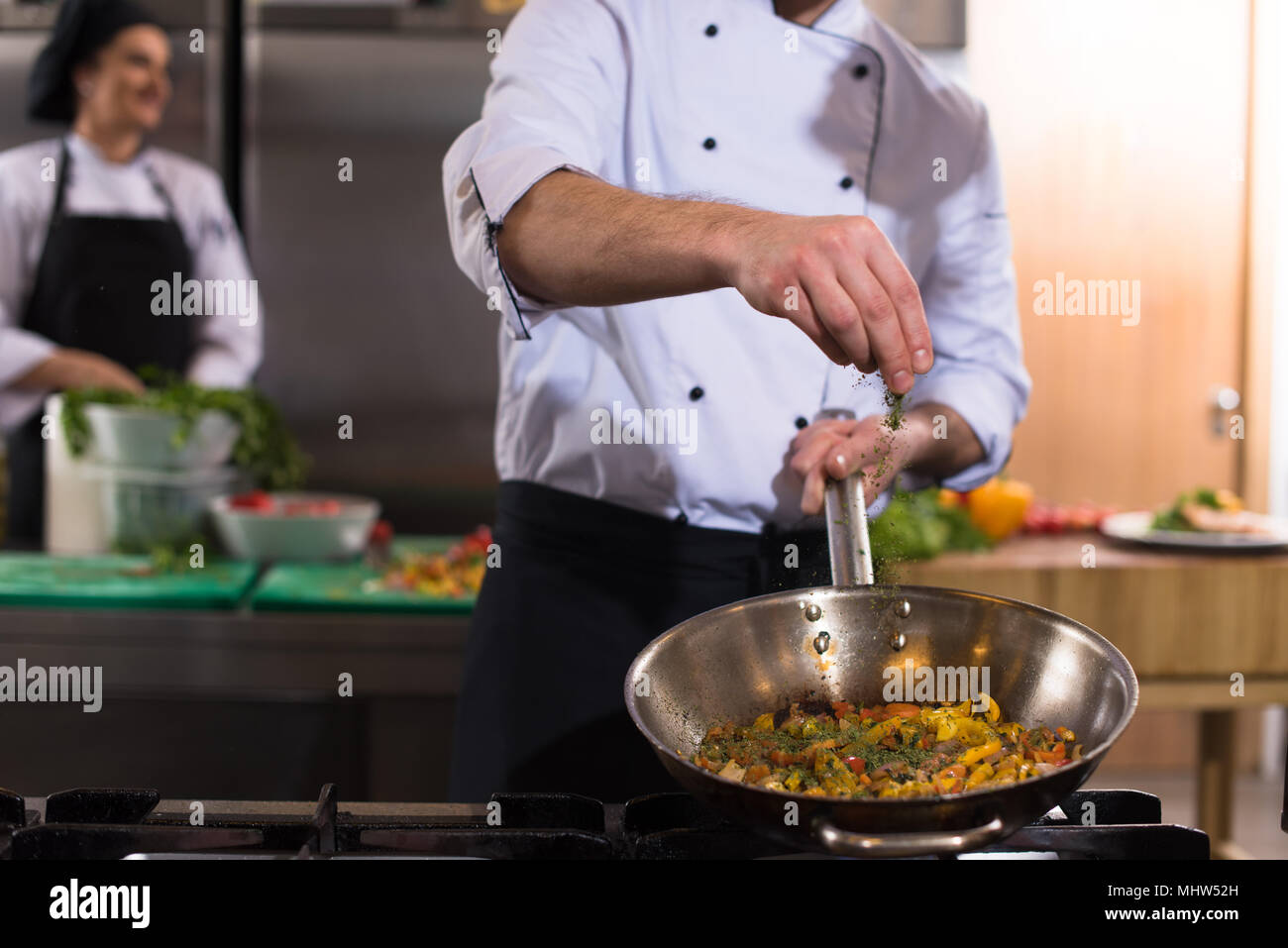 Young male chef putting spices on vegetables in wok at commercial ...