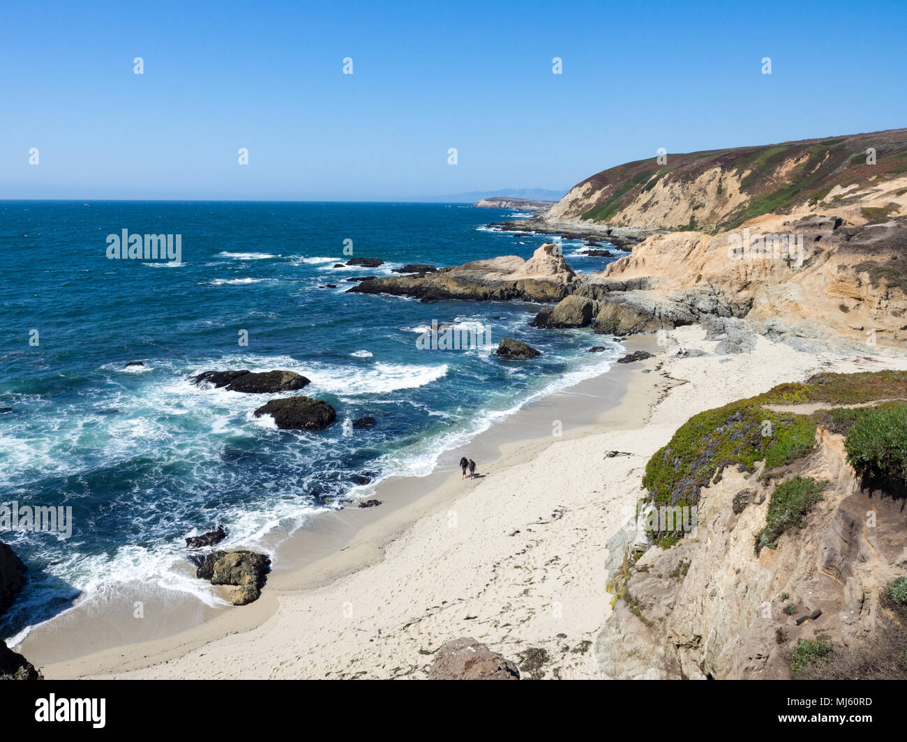 Barefoot hikers on beach below rocky cliffs and waves along shore in ...