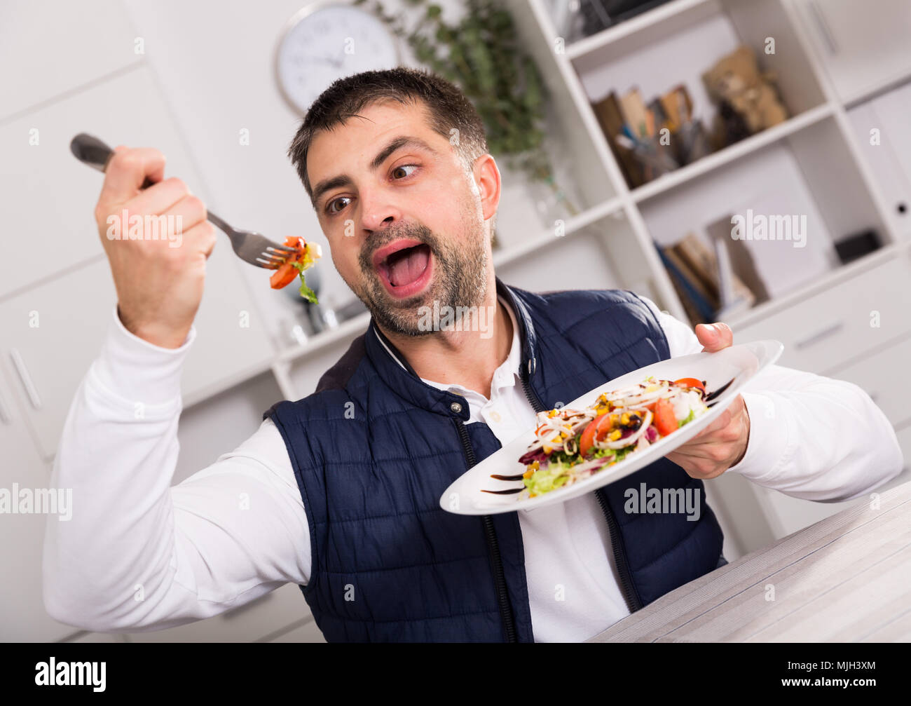 Positive young male holding fork and eating vegetable salad at table ...