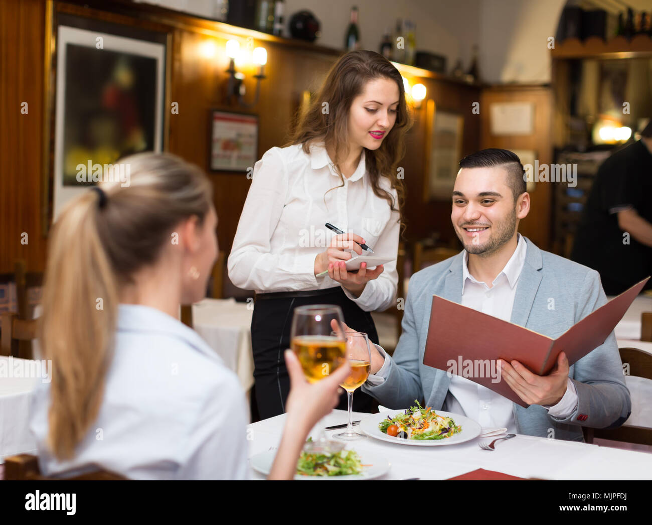 Young waitress offering tasty dishes to guests couple. Focus on man ...